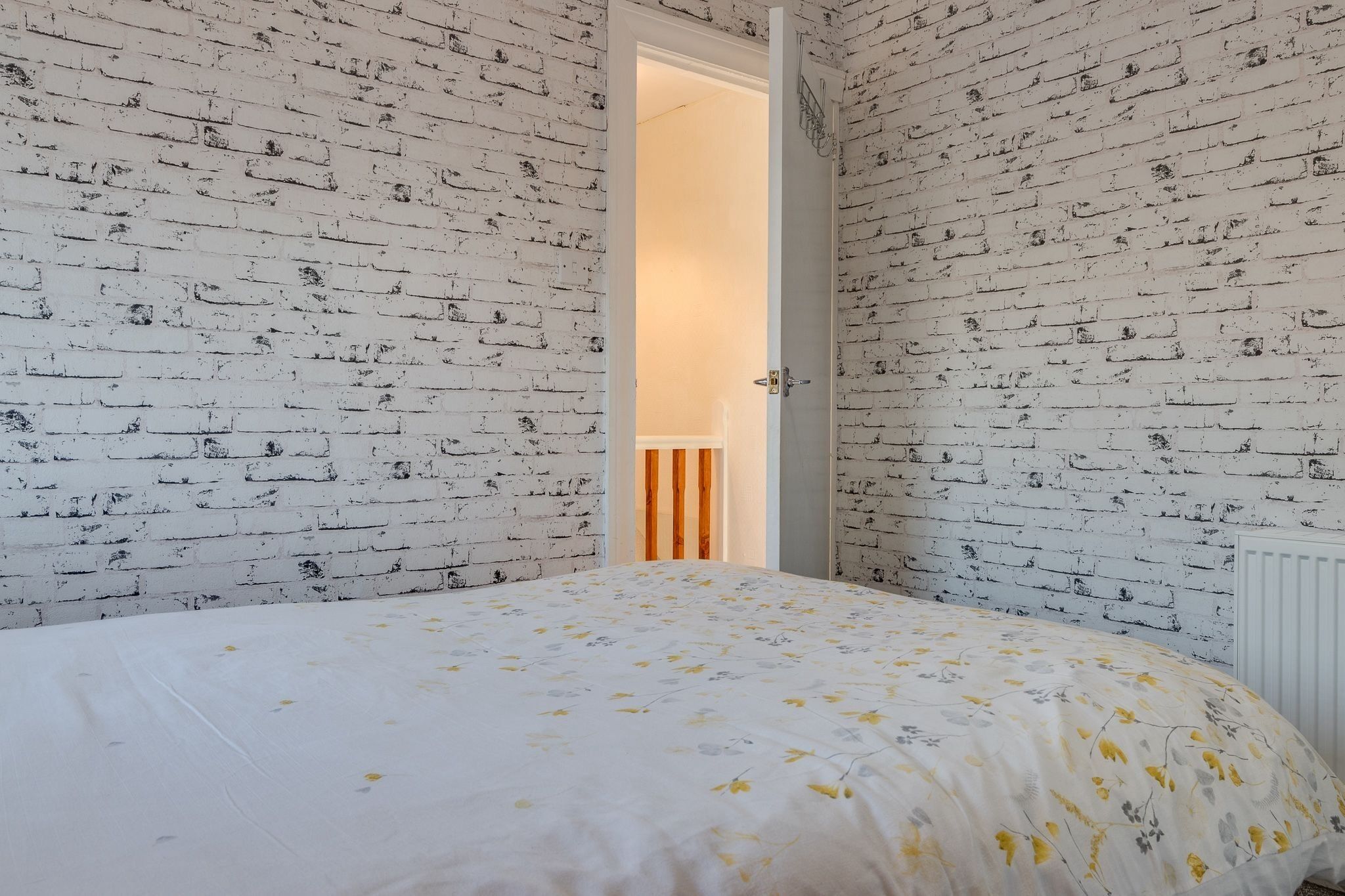 Quarto com parede em padrão tijolo, cama com colcha florida e porta que dá para outro cômodo.