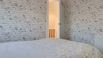 Quarto com parede em padrão tijolo, cama com colcha florida e porta que dá para outro cômodo.