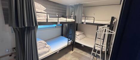 Quarto compacto com beliches, janela, cortinas e cama com colchão e almofadas.