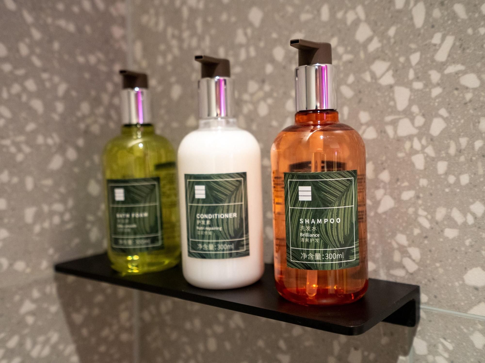 Três frascos de shampoo sobre uma prateleira preta em frente a uma parede texturizada.