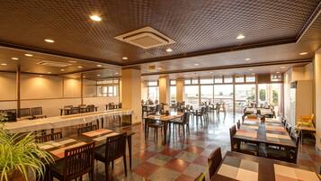 Um restaurante espaçoso com várias mesas e cadeiras, amplas janelas e teto com padrão decorativo.