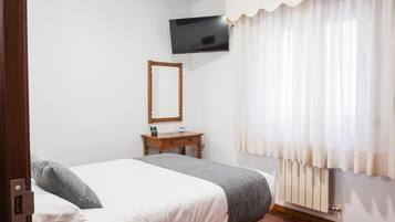 Quarto de hotel com cama, mesa de cabeceira de madeira, espelho, televisão na parede e janela com cortinas brancas.