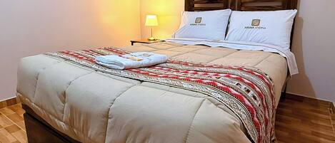 Uma cama bem arrumada com manta e almofadas estampadas, uma luminária de cabeceira e cabeceira de madeira.