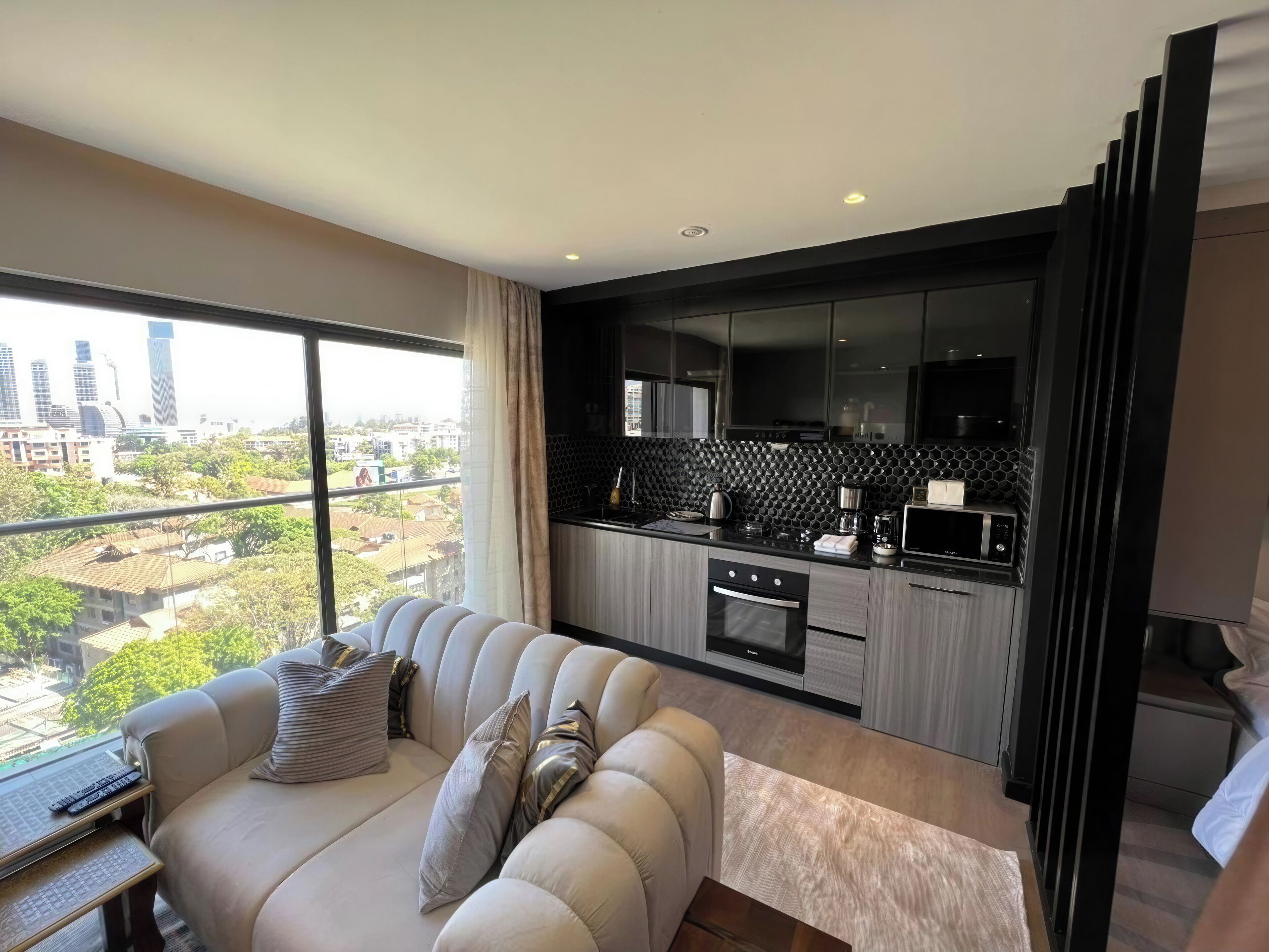 Apartamento moderno com sala de estar que inclui sofá e mesa de centro, cozinha com bancadas pretas e micro-ondas, e vista para a cidade através de grandes janelas.