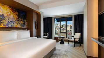 Quarto de hotel com uma cama grande, uma escrivaninha, uma cadeira, uma televisão e uma janela ampla com vista para a cidade.
