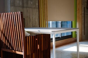 Uma cadeira de madeira com ripas verticais e uma mesa branca em um quarto com parede de concreto e janela com persianas.