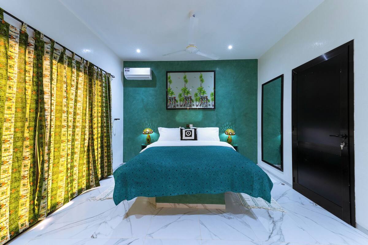 Quarto com cama, duas luminárias de cabeceira, ar-condicionado fixado na parede, um quadro e uma porta preta.