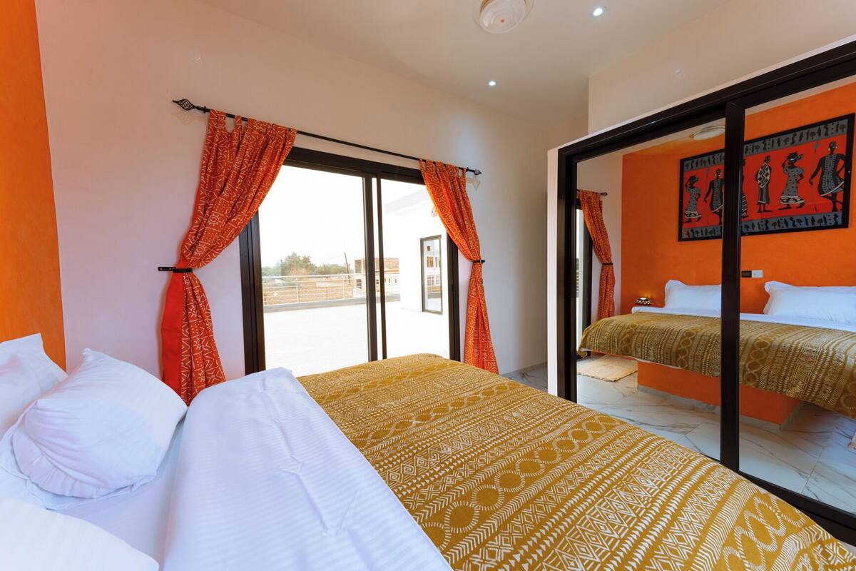 Quarto de hotel com duas camas, cortinas laranja, uma janela grande e um mural na parede.