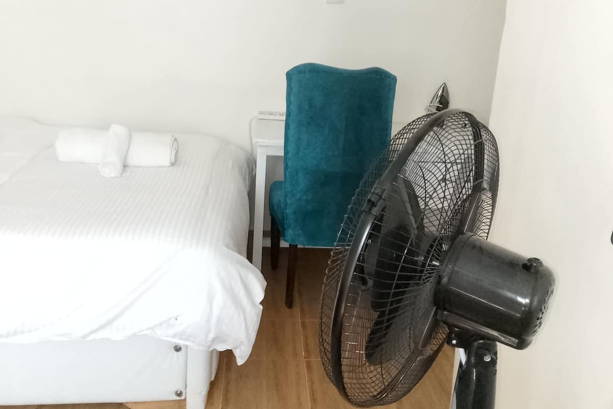 Quarto pequeno e bem organizado, com uma cama, uma cadeira, uma escrivaninha e um ventilador.