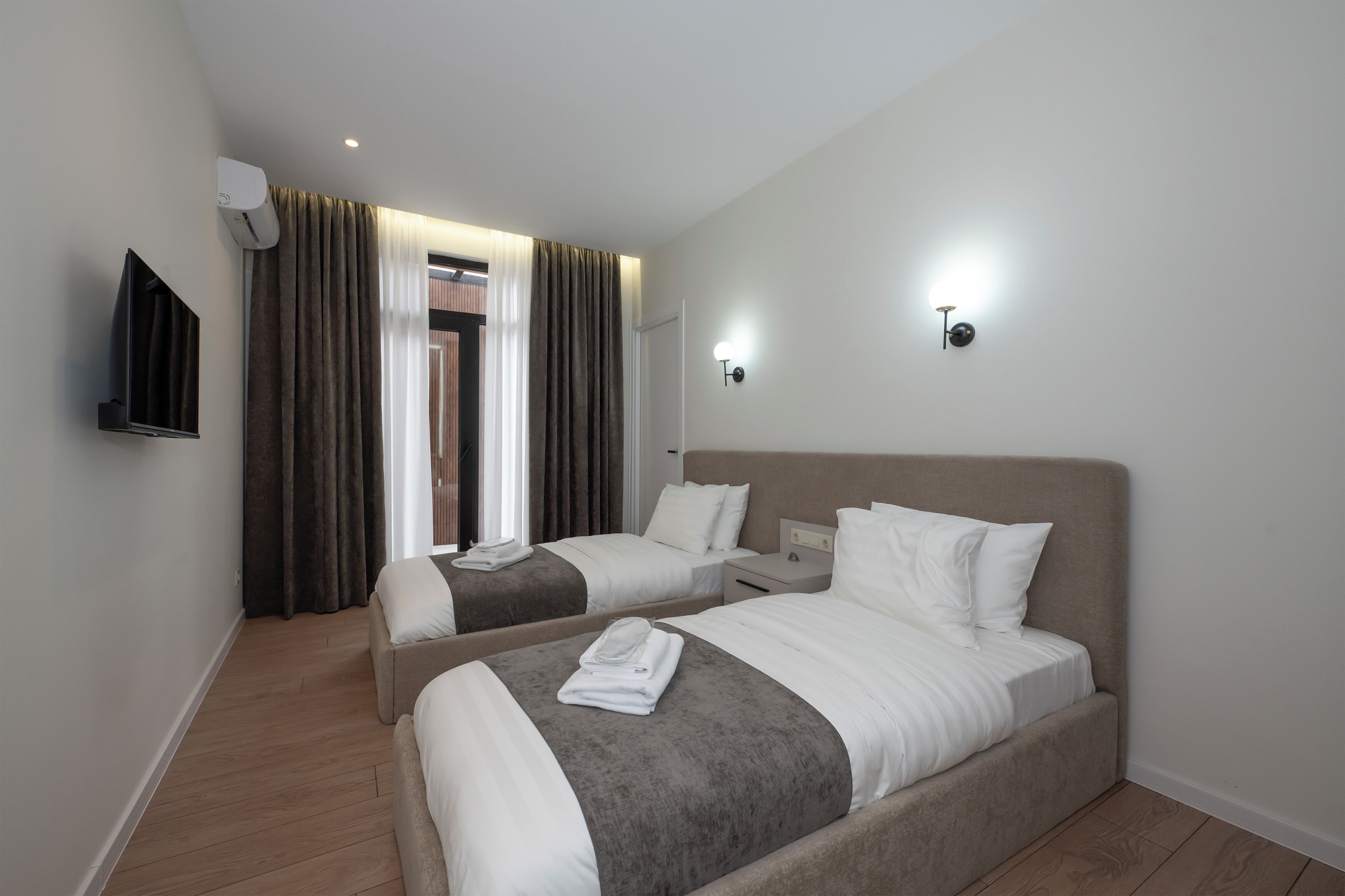 Quarto de hotel com duas camas, televisão, janela com cortinas e luminária fixada na parede.
