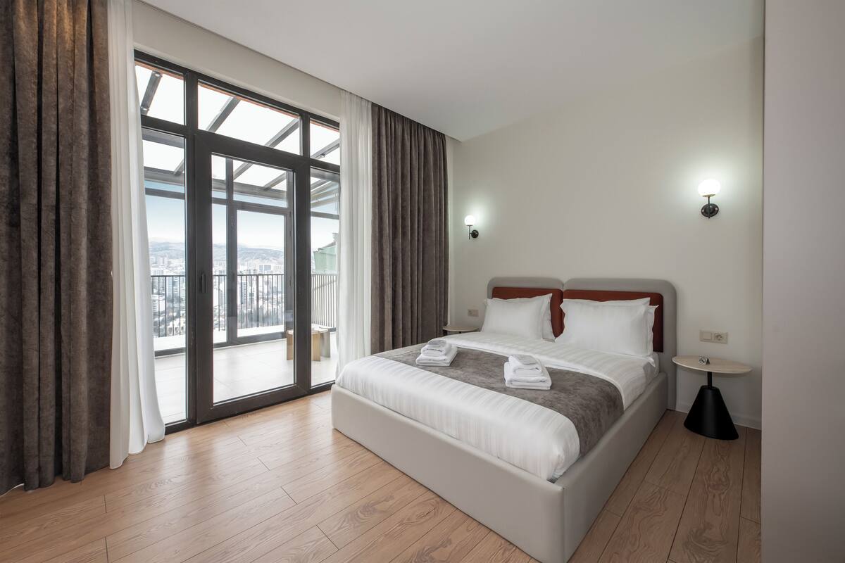 Quarto de hotel com uma cama grande, uma mesa redonda e vista para a cidade através de uma porta de vidro.