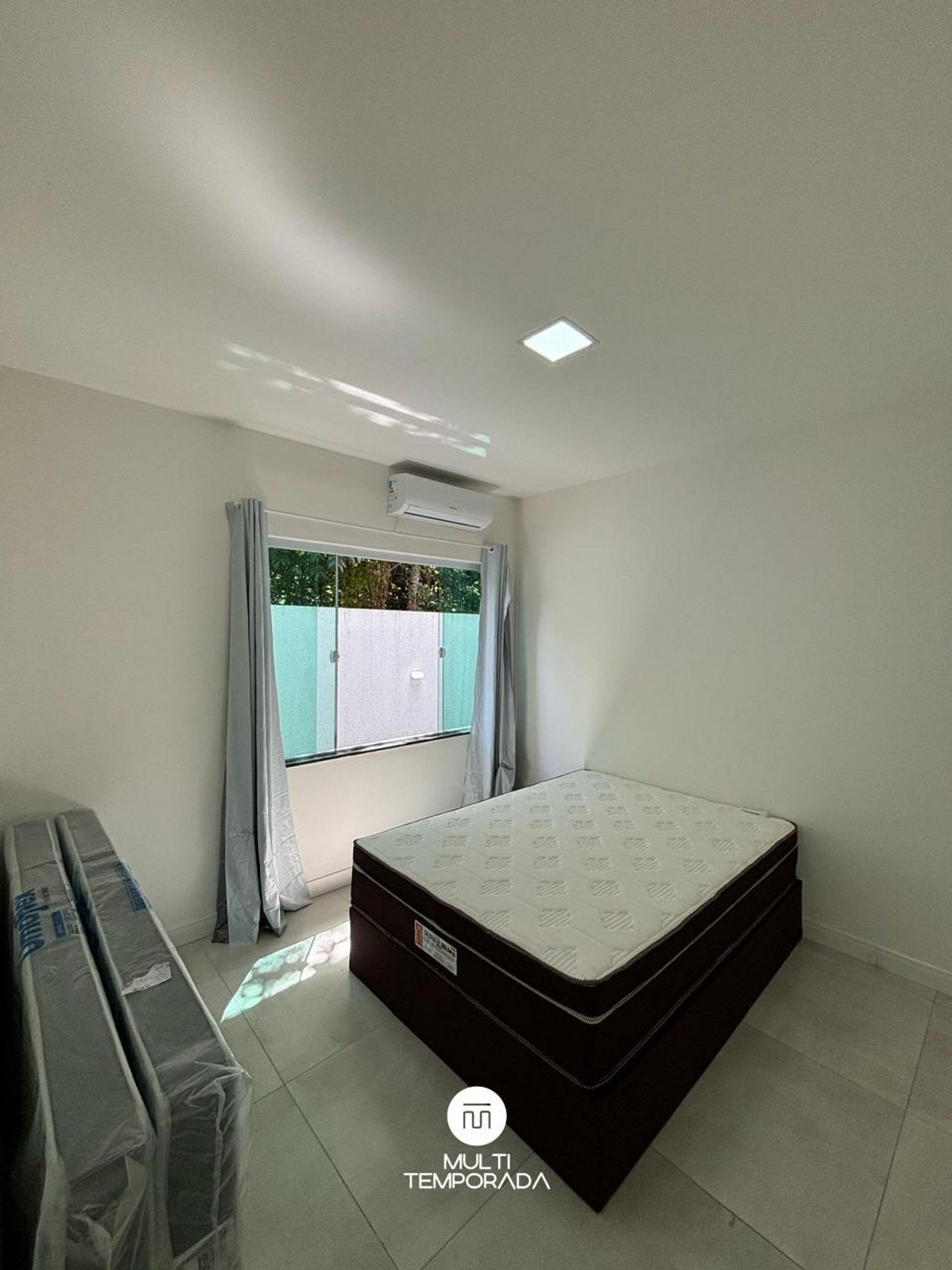 Quarto com cama grande, janela com cortinas, aparelho de ar condicionado e colchão no chão.
