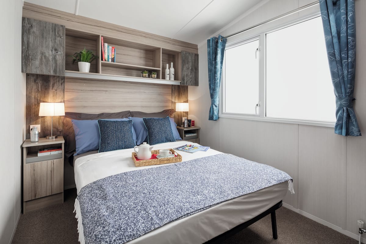 Um quarto bem arrumado com uma cama, mesas de cabeceira, estantes embutidas com livros e objetos decorativos, e uma janela com cortinas.
