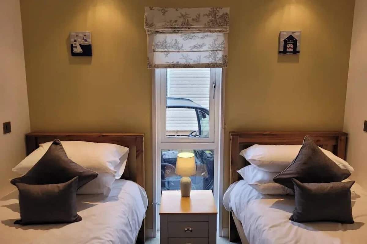 Quarto duplo com duas camas, uma mesinha de cabeceira, uma janela com vista e uma luminária.