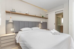 Quarto moderno com uma cama grande, mesas de cabeceira, uma prateleira flutuante com objetos decorativos e iluminação pendente.