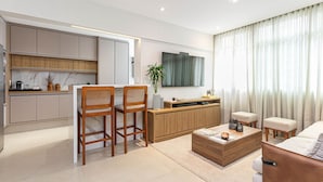 Uma sala de estar moderna com um sofá, mesa de centro e duas cadeiras de madeira. Há uma televisão montada na parede e uma área de cozinha com armários e bancada.