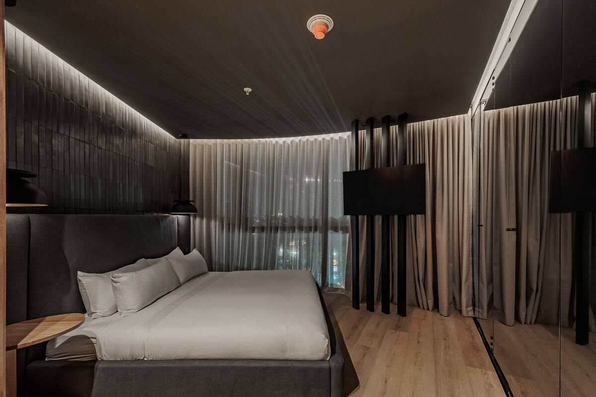 Quarto moderno com cama grande, televisão de tela plana e vista para a cidade através de cortinas translúcidas.