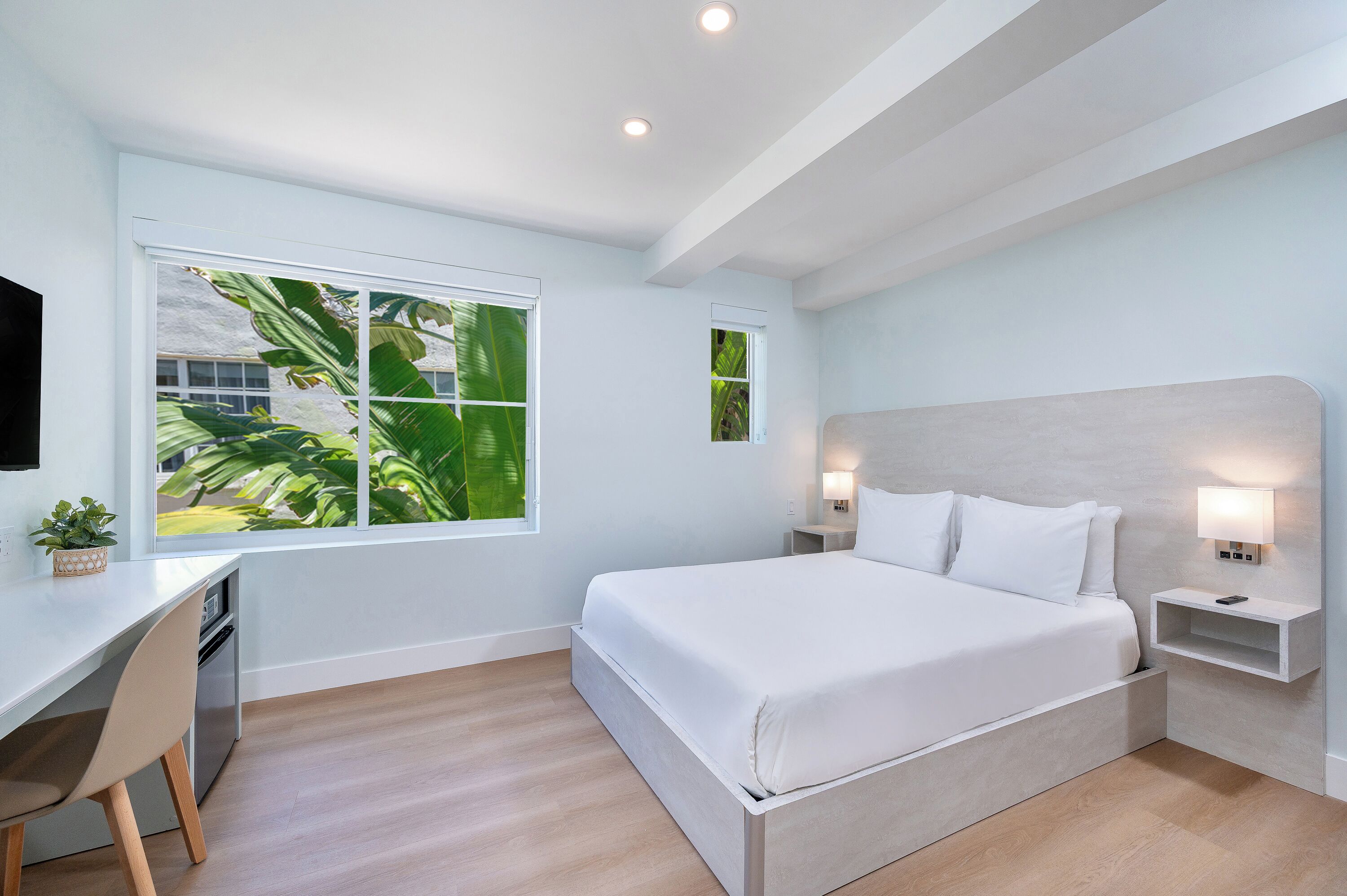 Quarto moderno com cama, mesas de cabeceira, escrivaninha e uma janela grande com vista para o verde.