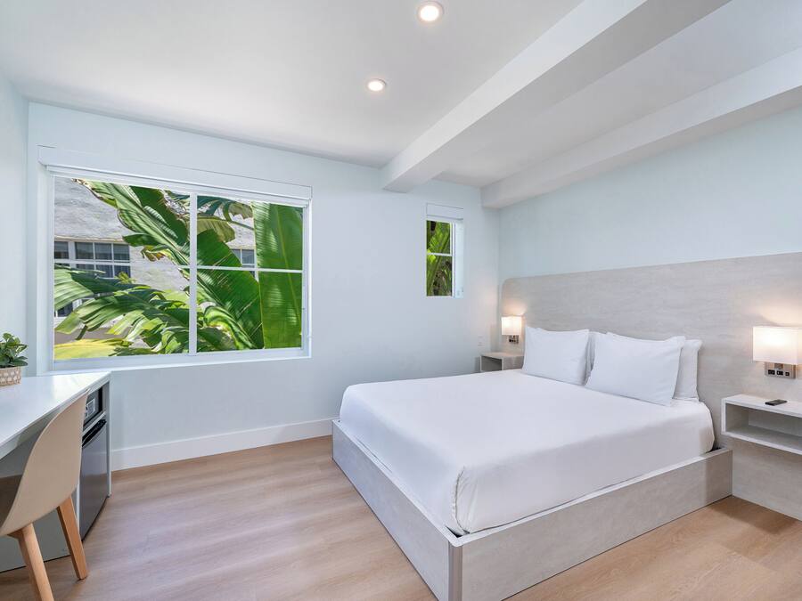 Quarto moderno com cama, mesas de cabeceira, escrivaninha e uma janela grande com vista para o verde.