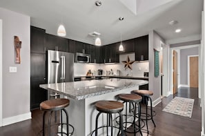 Cozinha moderna com armários escuros, eletrodomésticos de aço inoxidável, uma ilha grande com banquetas, iluminação pendente e um objeto decorativo em formato de estrela.