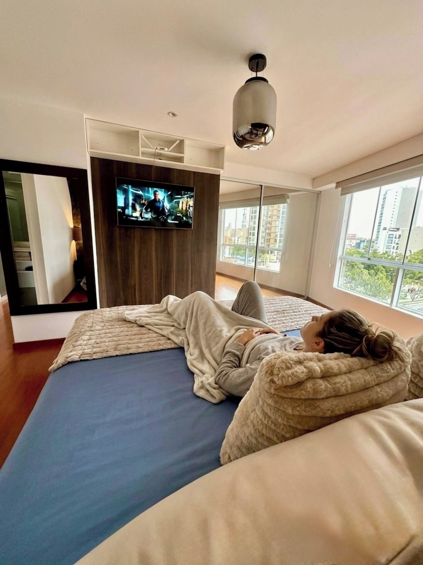 Quarto moderno com cama grande, televisão de tela plana fixada em um painel de madeira e vista para a cidade por grandes janelas.