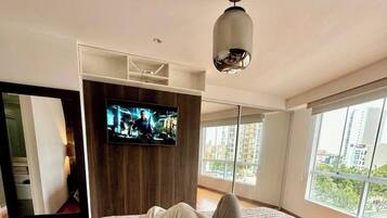 Quarto moderno com cama grande, televisão de tela plana fixada em um painel de madeira e vista para a cidade por grandes janelas.