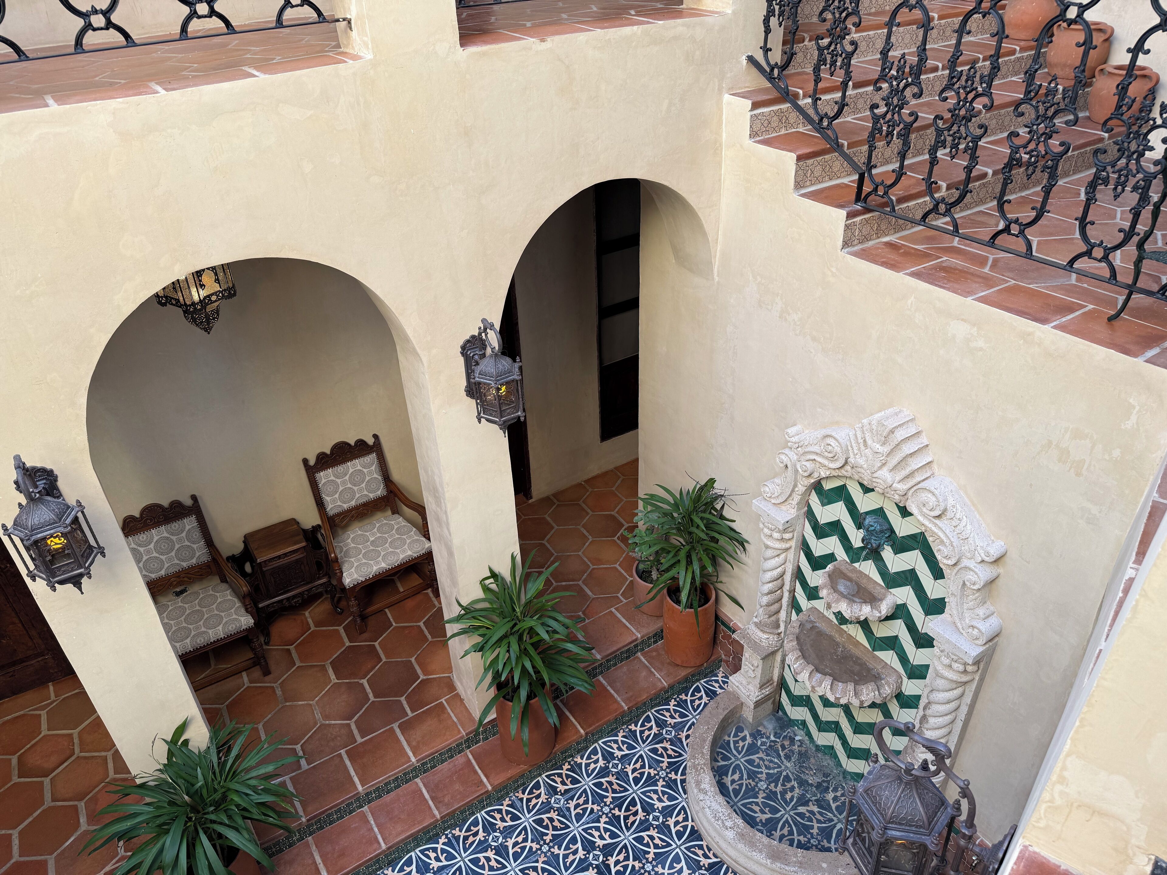 Um pátio com piso de azulejos, duas cadeiras de madeira, plantas em vasos, uma fonte decorativa e grades de ferro forjado.
