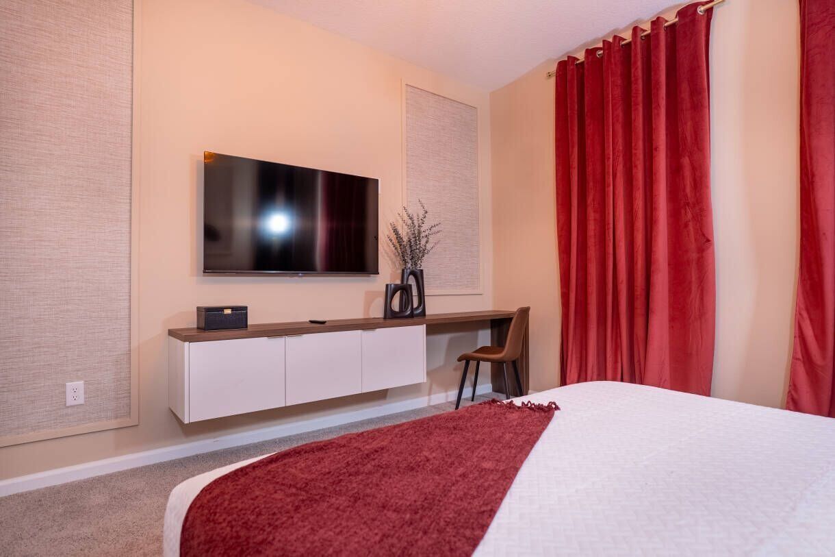 Quarto de hotel moderno com uma cama grande, uma TV de tela plana na parede, uma escrivaninha com cadeira e uma cortina vermelha.