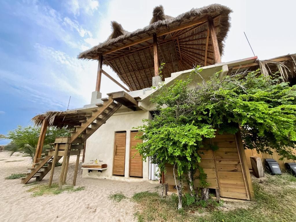Uma casa de praia com escada para um deck de madeira, telhado de palha e um pequeno jardim.