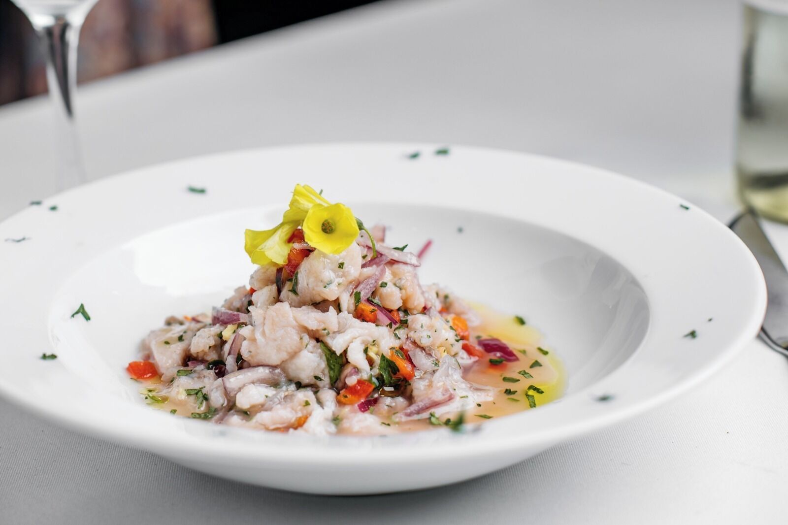 Prato branco com prato de frutos do mar, decorado com ervas e uma flor amarela, sobre uma toalha de mesa branca.