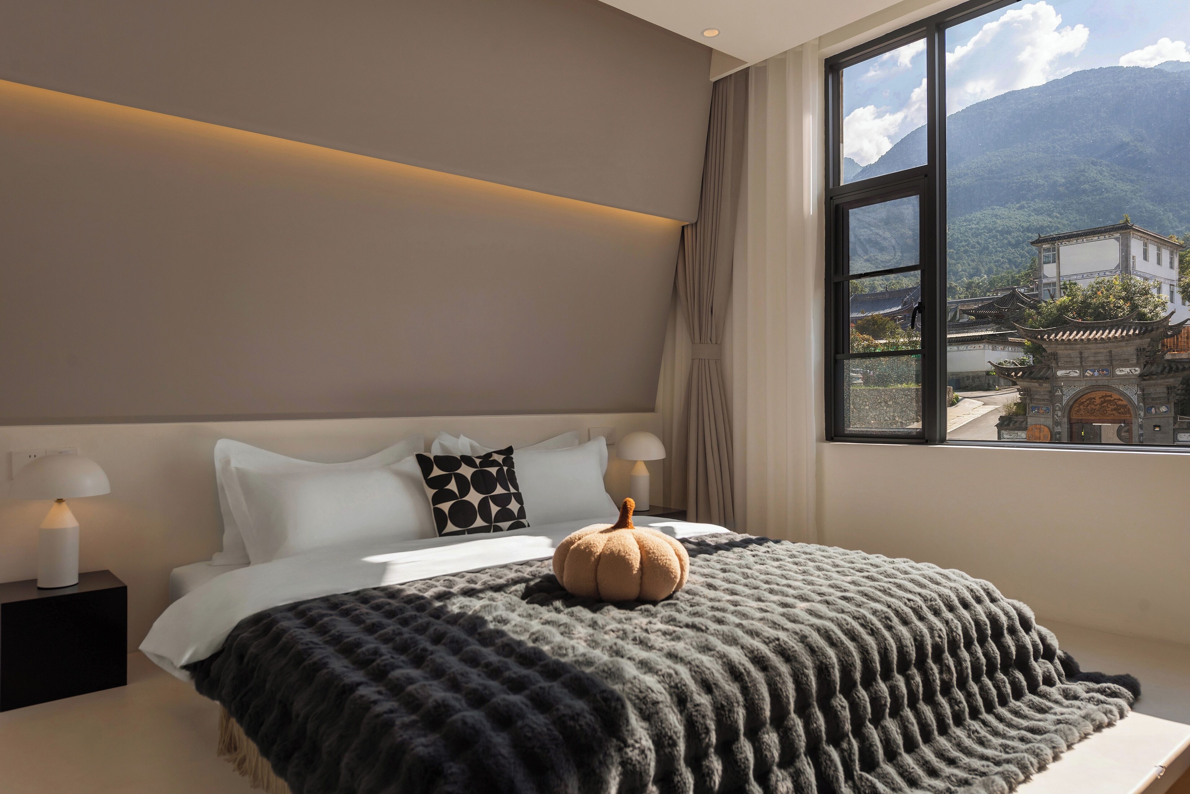 Quarto com uma cama grande, duas luminárias de cabeceira, um travesseiro decorativo, uma abóbora macia e vista da arquitetura tradicional pela janela.