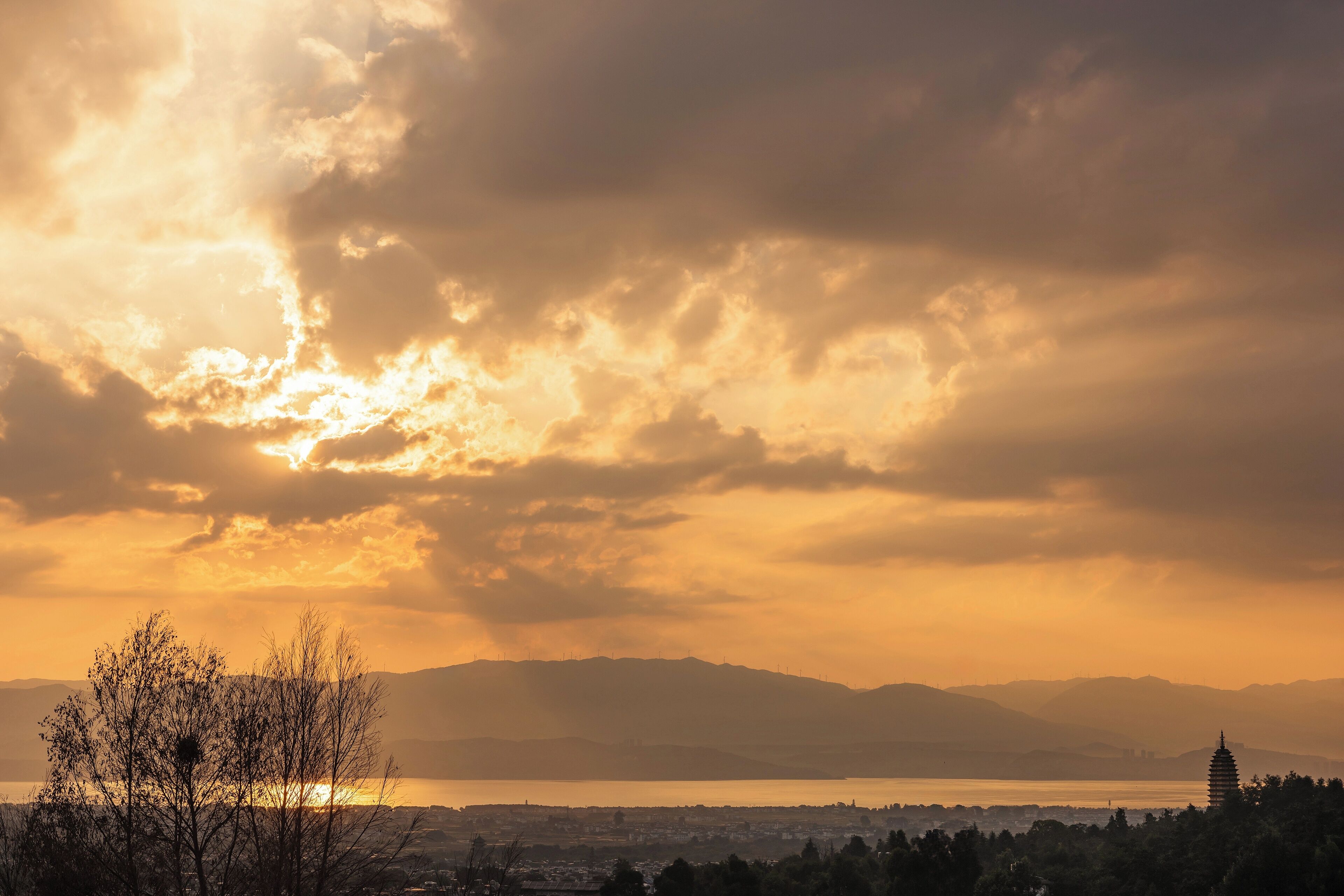 Vista panorâmica de uma paisagem urbana com uma pagoda imponente, um corpo d'água e montanhas ao longe sob um céu ao pôr do sol.