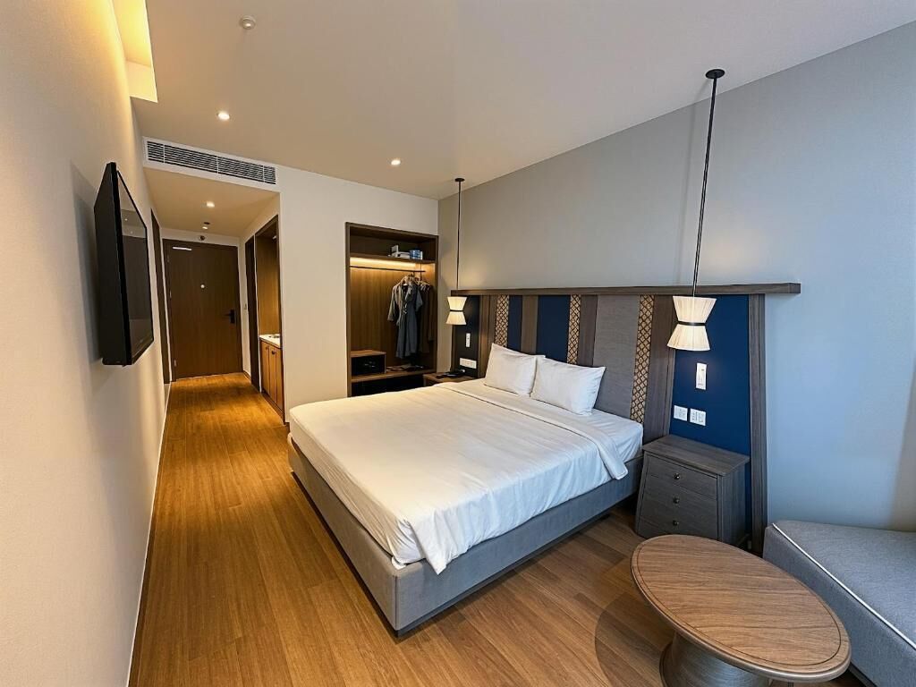 Quarto de hotel moderno com uma cama grande, mesa de cabeceira, cadeira, guarda-roupa e televisão presa na parede.