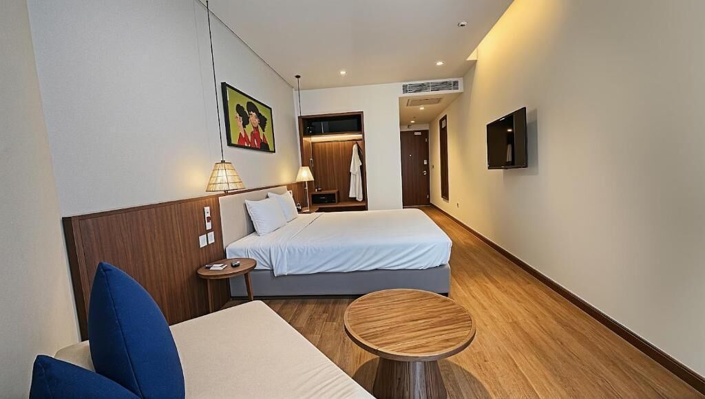 Quarto de hotel moderno com cama, mesa de cabeceira, abajur e uma mesa redonda de madeira.