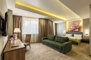 Quarto de hotel com uma cama, um sofá verde, uma escrivaninha de madeira, uma cadeira e uma televisão.