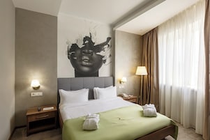 Quarto de hotel com uma cama grande, duas mesas de cabeceira, um criado-mudo, uma luminária fixada na parede e uma janela grande com cortinas.