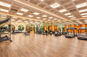Academia com diversos equipamentos de exercícios, piso de madeira e um espelho grande na parede.