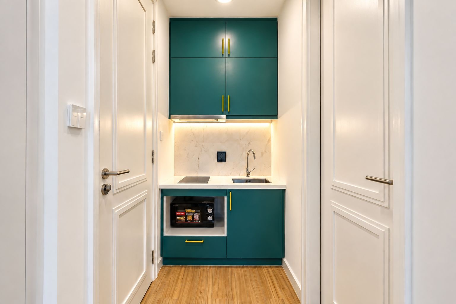 Corredor estreito que dá para uma cozinha com armários azul-turquesa, forno embutido e painel de fundo de mármore.