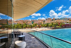 Um hotel na praia com área de piscina, mesas ao ar livre e vista para o mar e edifícios coloridos ao fundo.
