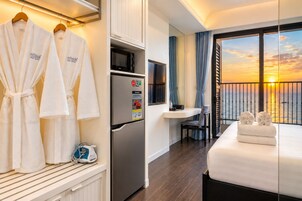 Quarto de hotel com uma cama, minibar, geladeira, televisão e vista para o pôr do sol sobre a água.