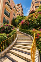 Escadaria com azulejos decorados, rodeada por vegetação exuberante e flores vibrantes, conduzindo a um edifício com varandas.