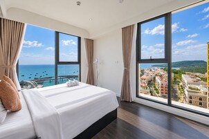 Quarto de hotel com uma cama grande, duas janelas e vista para o mar e a cidade.