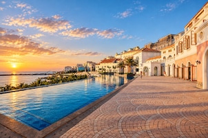 Piscina com vista para o pôr do sol sobre o mar, cercada por edifícios com janelas em arco e um calçadão pavimentado.