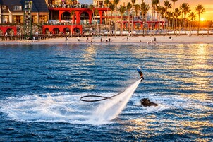 Um jet ski realiza uma manobra com uma pessoa a bordo, com água espirrando e uma praia com palmeiras e edifícios ao fundo.