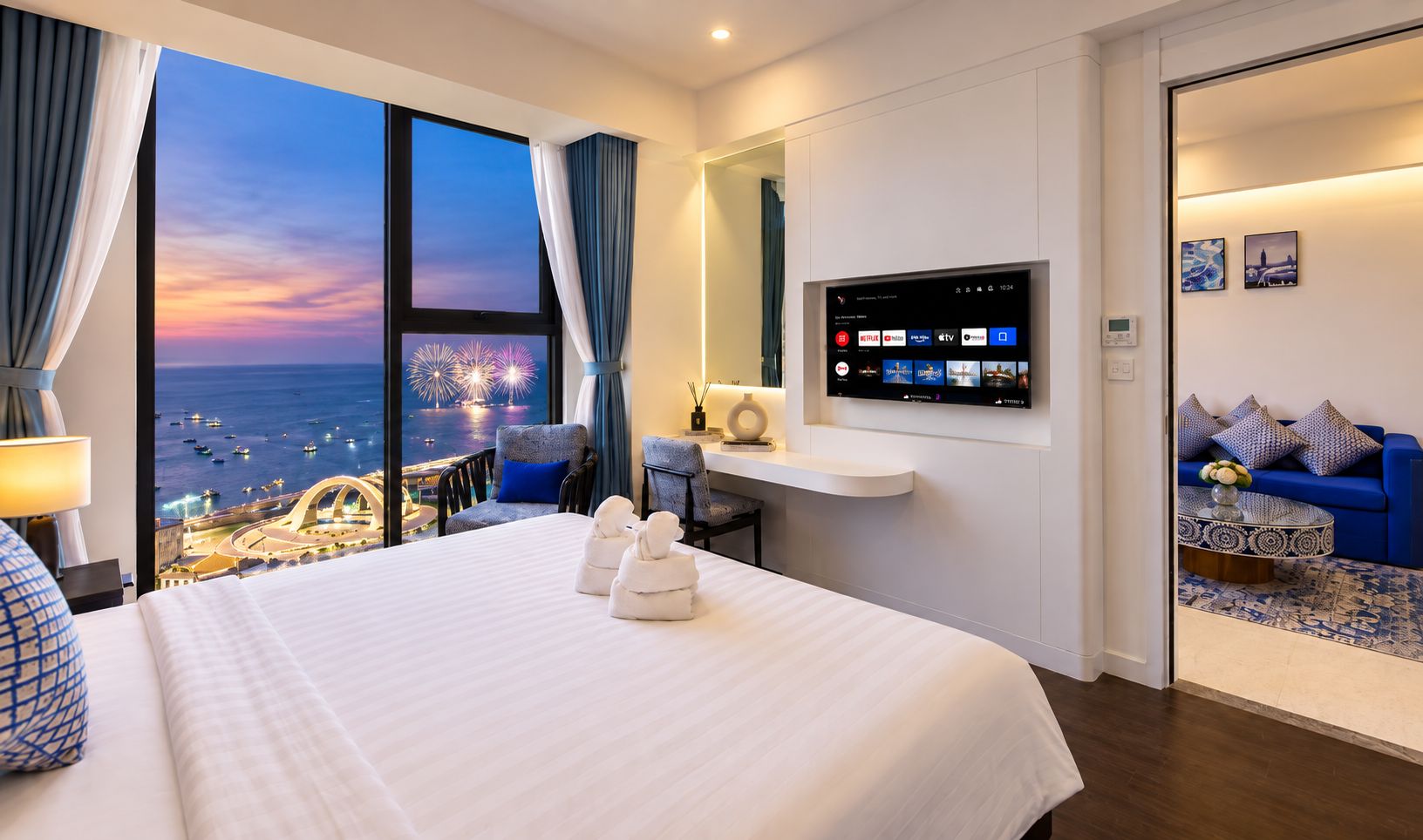 Quarto de hotel com cama, televisão na parede, área de estar com sofá e poltronas, e vista para o mar e fogos de artifício.