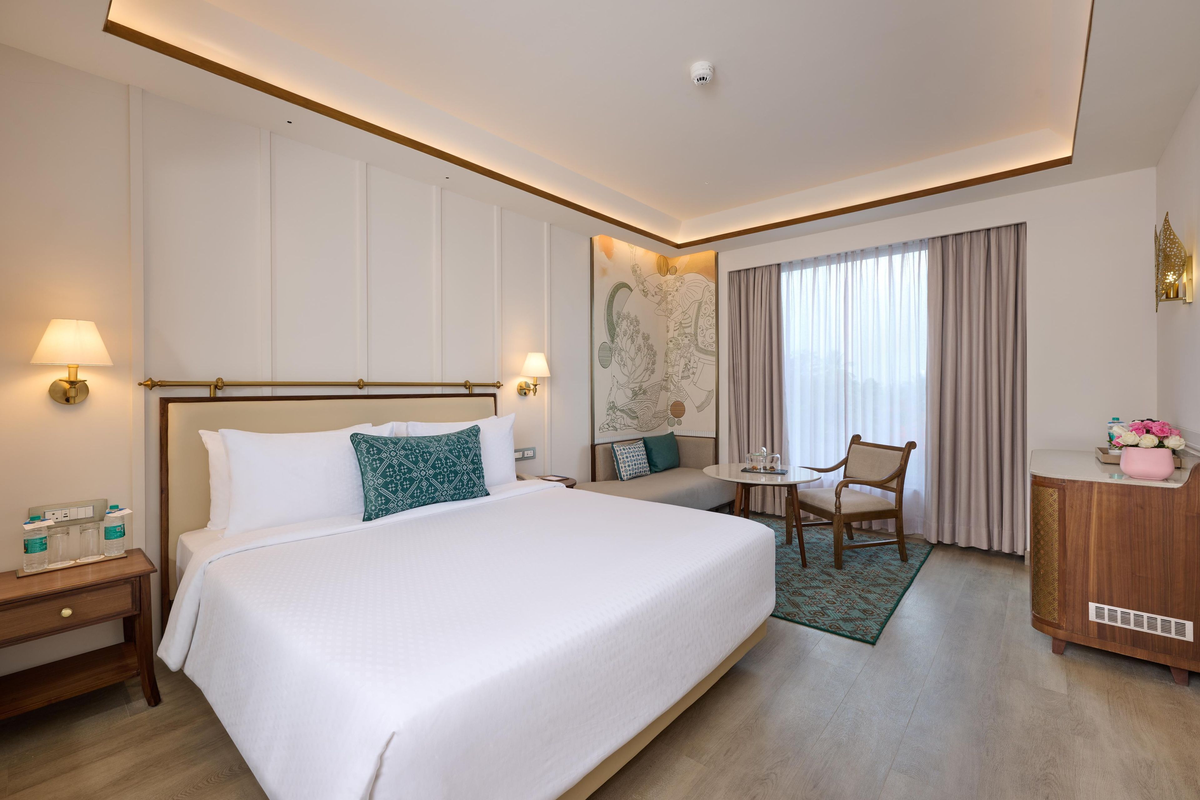 Quarto de hotel com uma cama grande, uma escrivaninha, uma cadeira e uma mesinha. Há uma janela com cortinas e uma parede com um mural decorativo.
