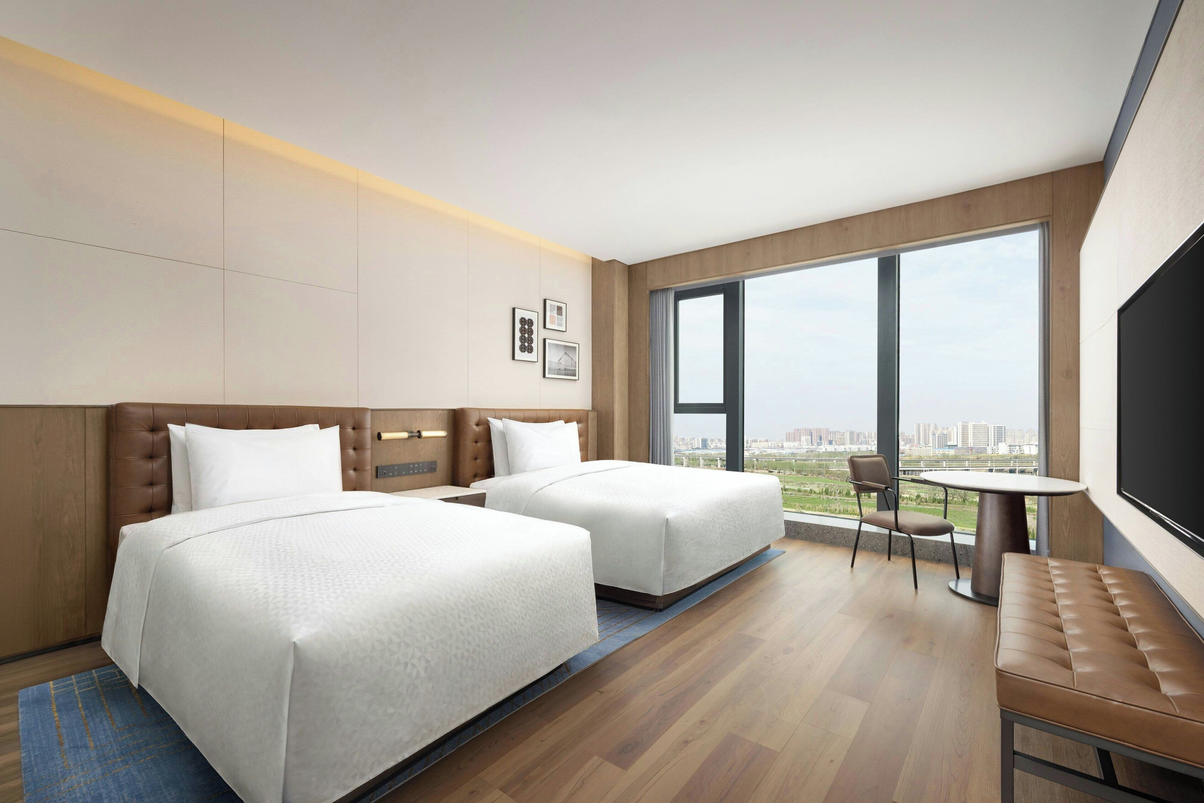 Quarto de hotel com duas camas, área de estar e uma janela grande com vista para a cidade.