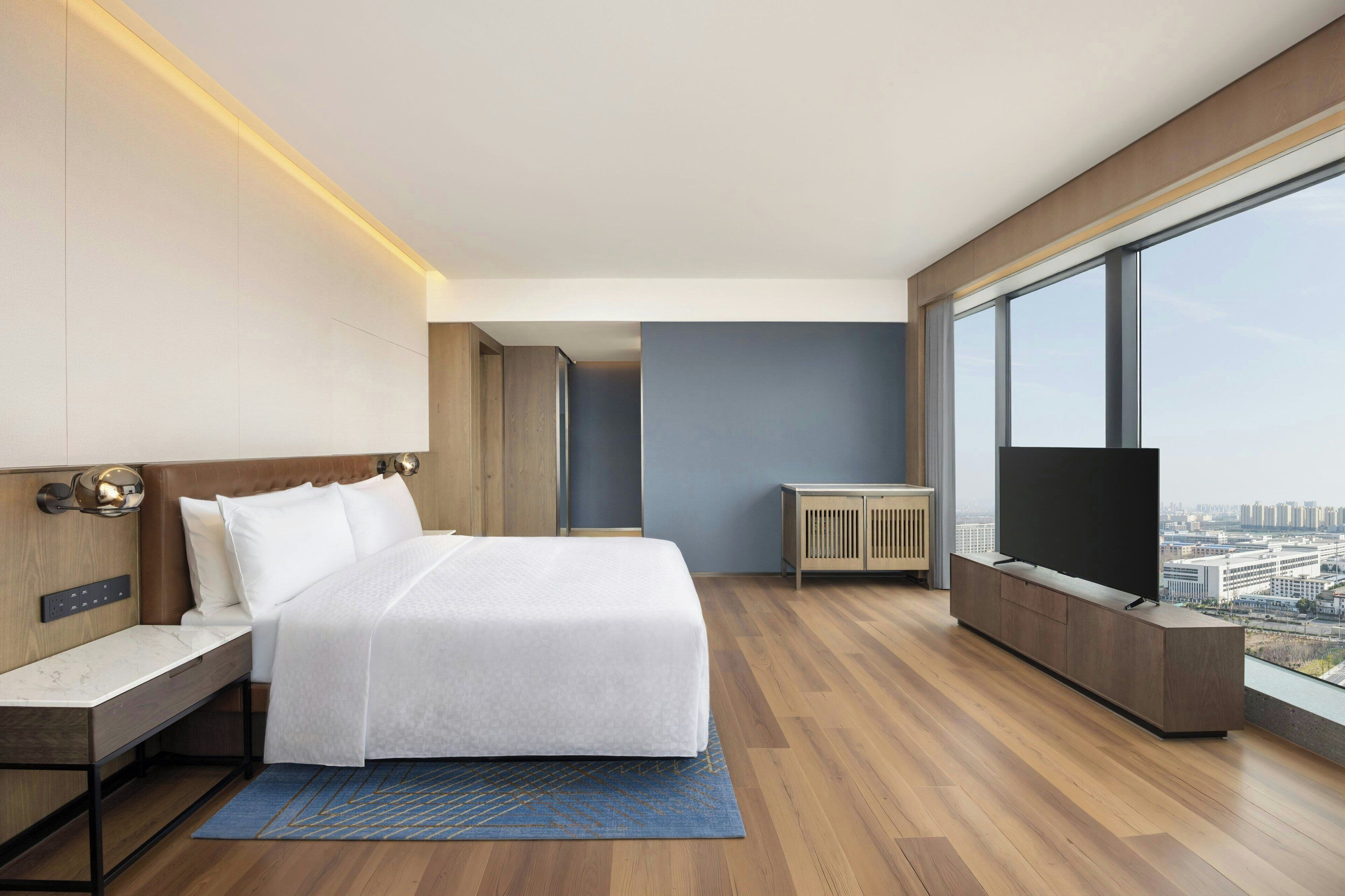 Quarto de hotel moderno com uma cama grande, mesa de cabeceira, televisão e vista da cidade através de amplas janelas.