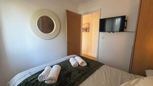 Cama bem arrumada com toalhas enroladas, um espelho redondo, televisão na parede e uma porta que dá para outro quarto.