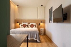 Quarto com cama, mesas de cabeceira, criado-mudo, televisão fixada na parede e cabeceira de madeira.